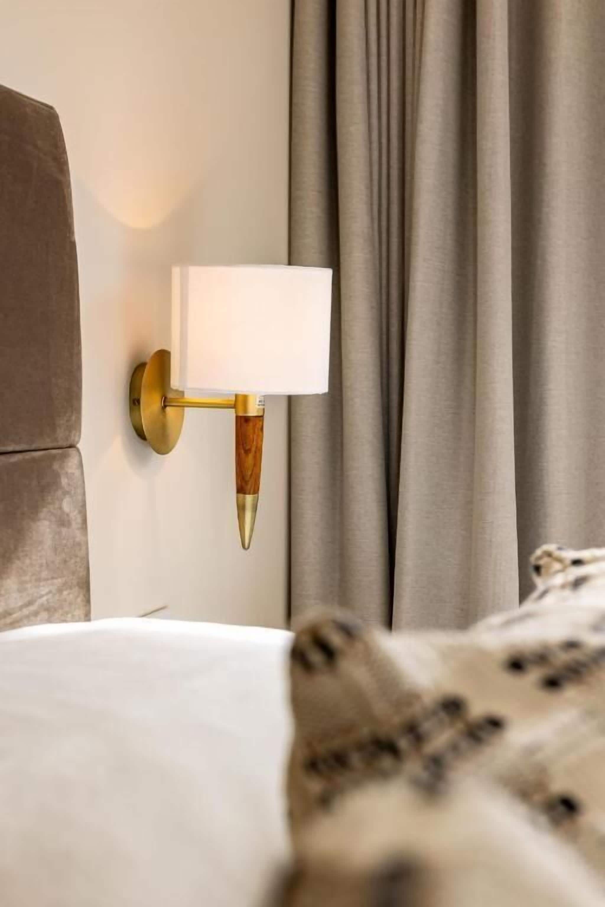 Uma cama com cabeceira branca, luminária fixada na parede com detalhe em madeira e cortinas cinzas.