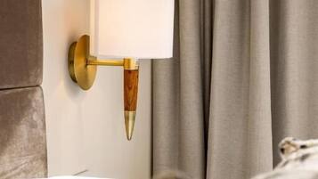 Uma cama com cabeceira branca, luminária fixada na parede com detalhe em madeira e cortinas cinzas.
