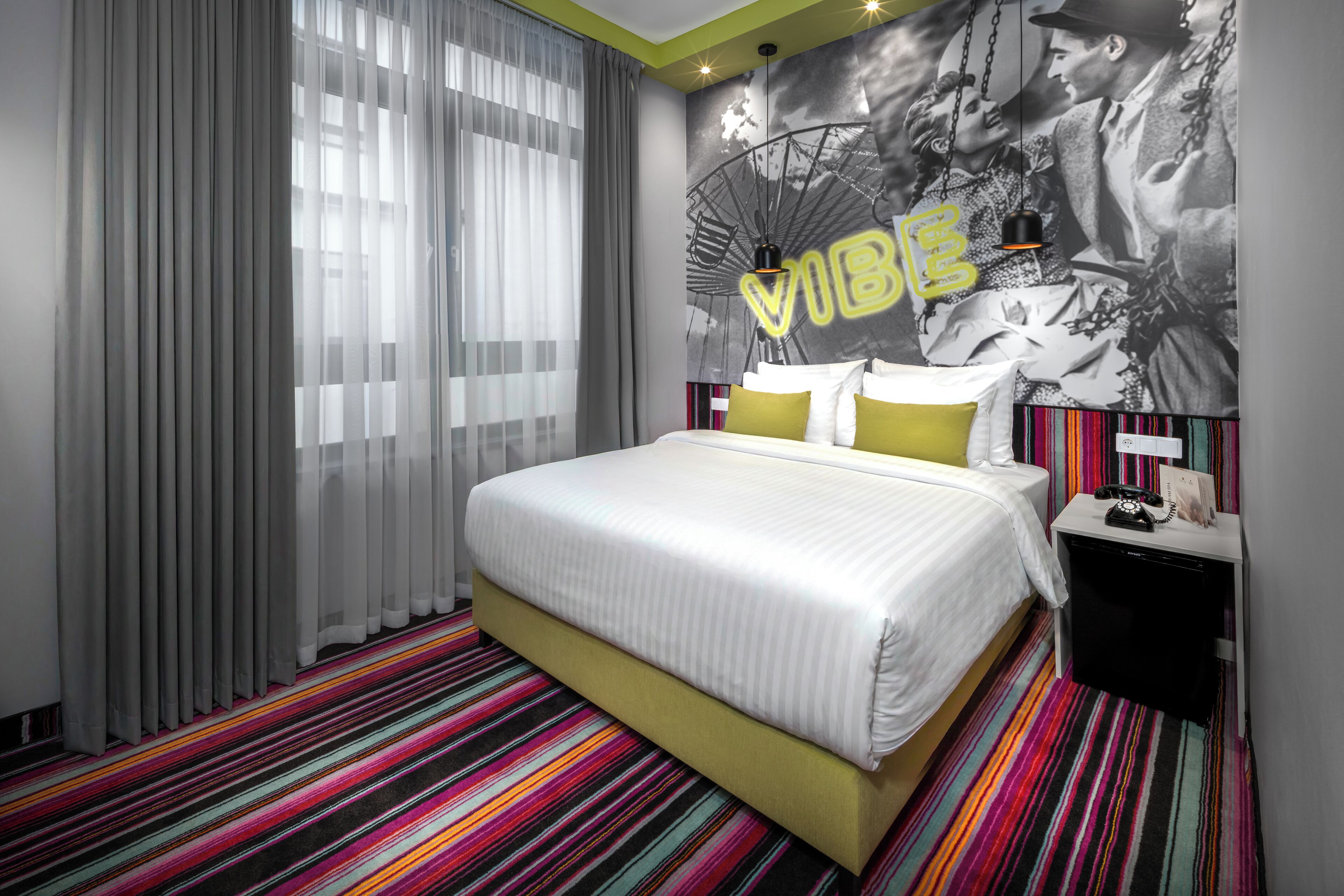 Quarto de hotel moderno com uma cama grande, carpete listrado colorido, luminária fixada na parede e um letreiro com a palavra 'VIBE'.