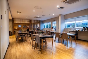 Uma sala de jantar moderna com piso de madeira, mesas e cadeiras. Grandes janelas oferecem luz natural e vista para o exterior.