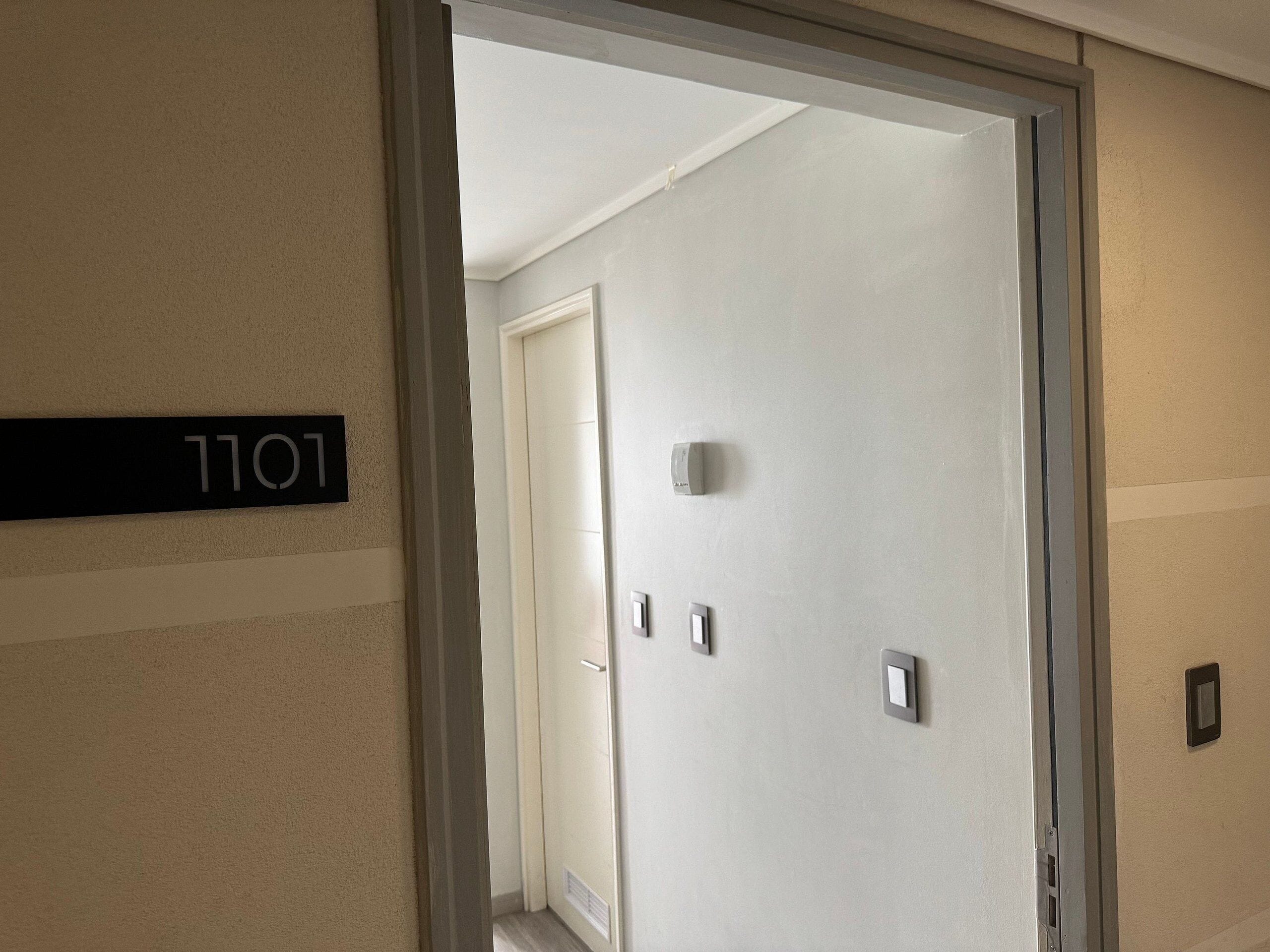 Um corredor com uma porta que dá para um quarto, um interruptor de luz fixado na parede e um interfone.