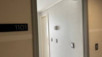 Um corredor com uma porta que dá para um quarto, um interruptor de luz fixado na parede e um interfone.