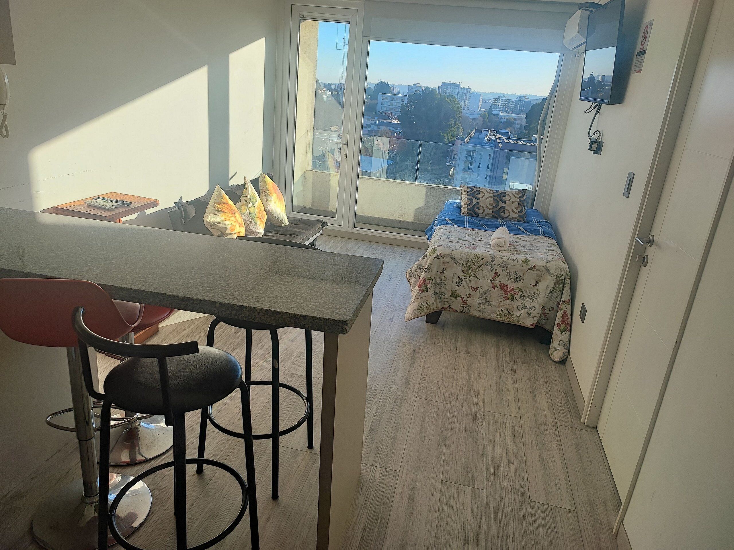 Quarto pequeno com cama, mesa e cadeira. Tem uma janela com vista para edifícios.