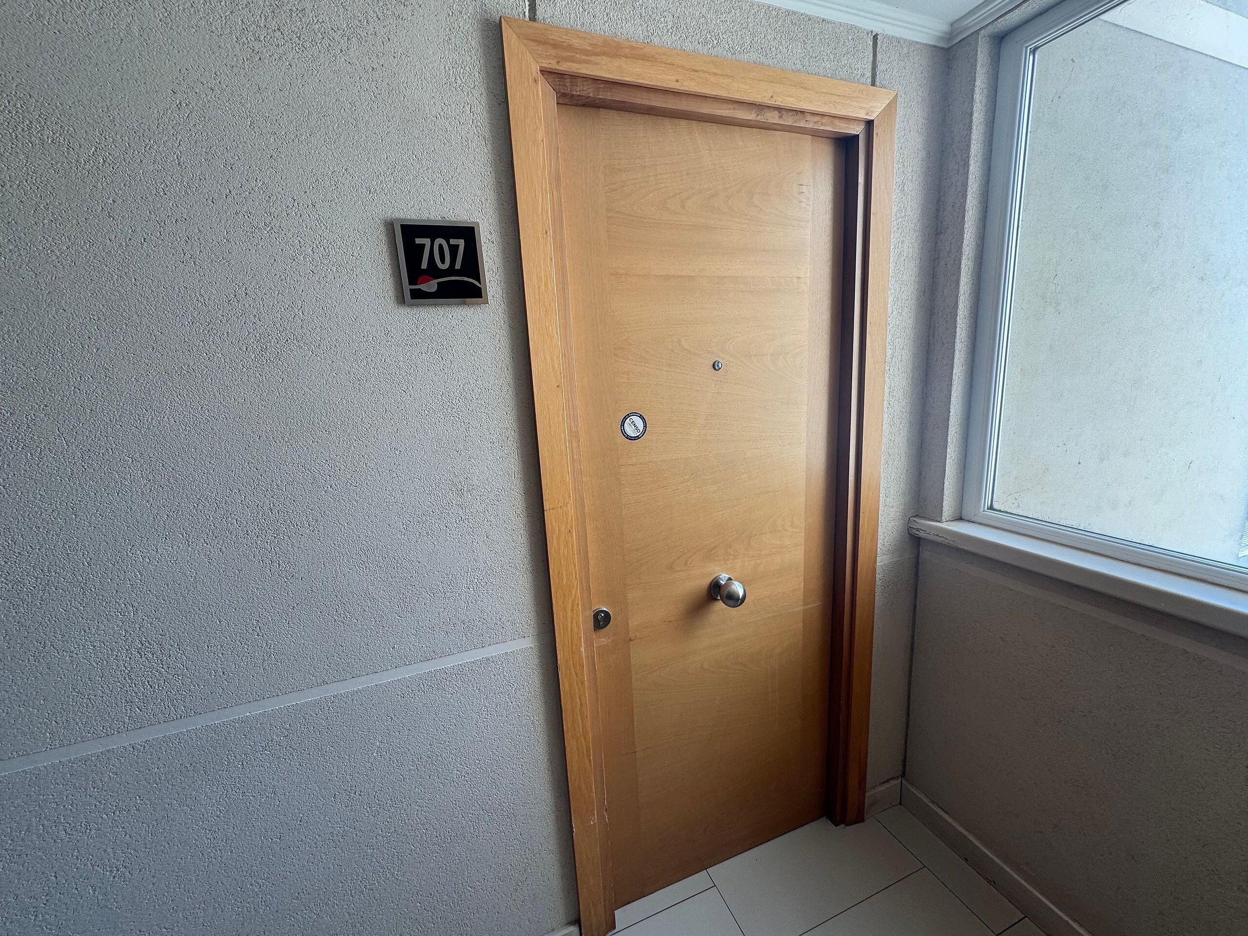 Uma porta de madeira com placa indicando o quarto 707, uma janela à direita e uma parede texturizada.