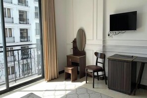 Quarto com uma janela grande, toucador com espelho, televisão na parede e uma pequena mesa.