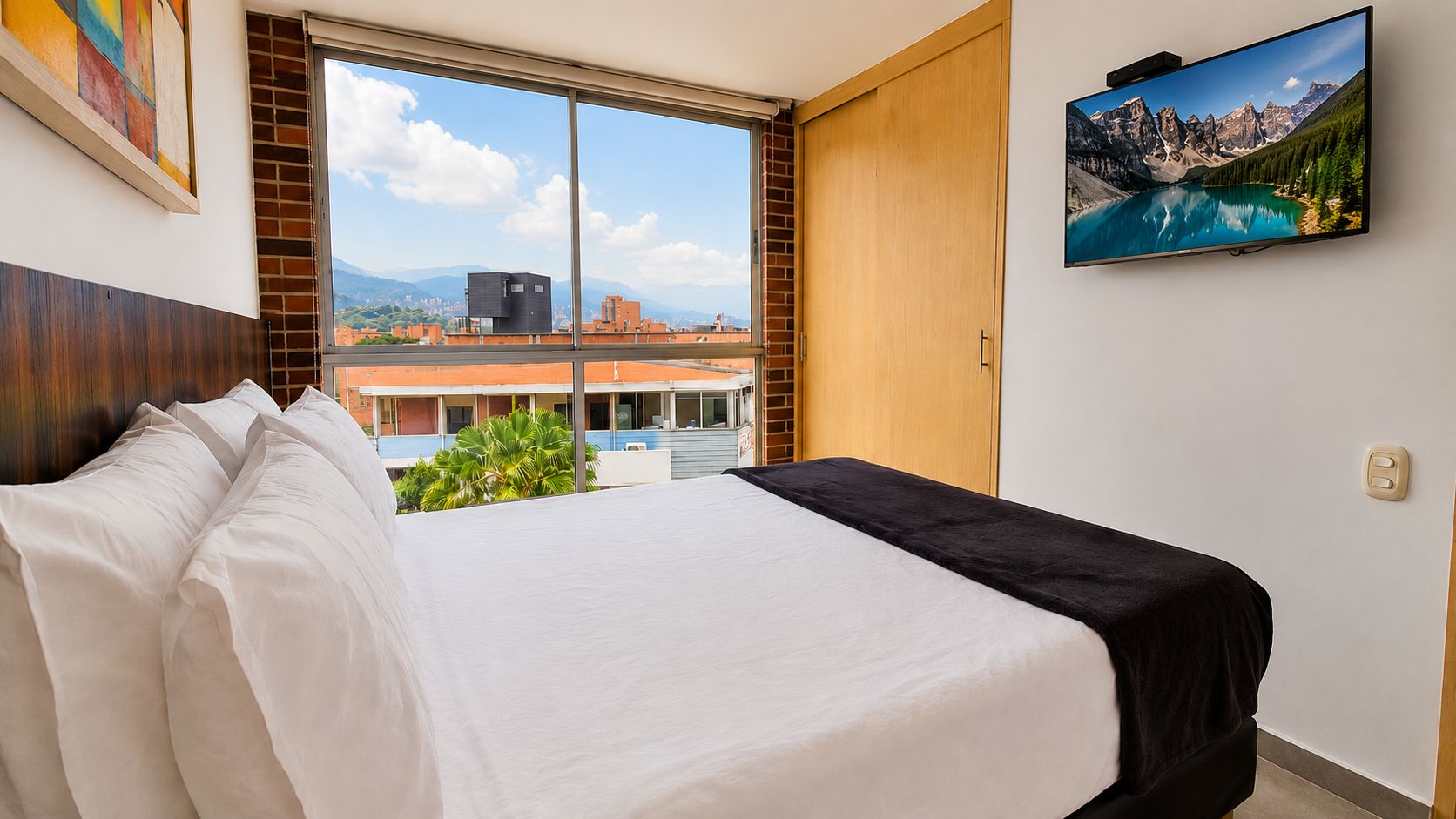 Quarto de hotel com cama, janela com vista para montanhas e um lago, e um quadro na parede.