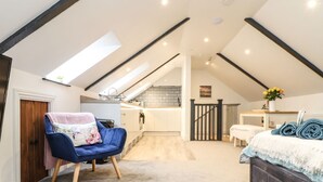 Um quarto amplo no estilo loft, com claraboia, uma cadeira azul aconchegante, uma cama com toalhas dobradas e uma cozinha ao fundo.
