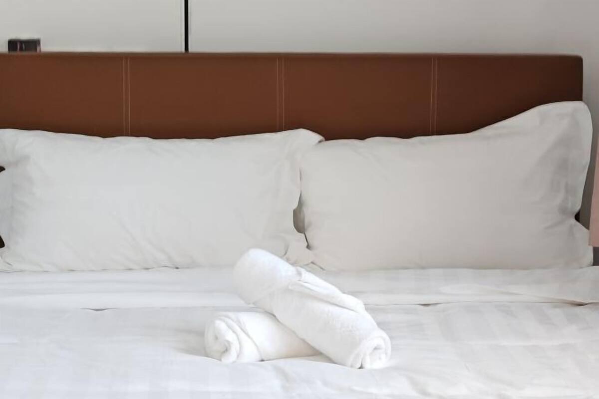 Cama bem arrumada com lençóis e almofadas brancos, uma toalha branca dobrada e cabeceira marrom.