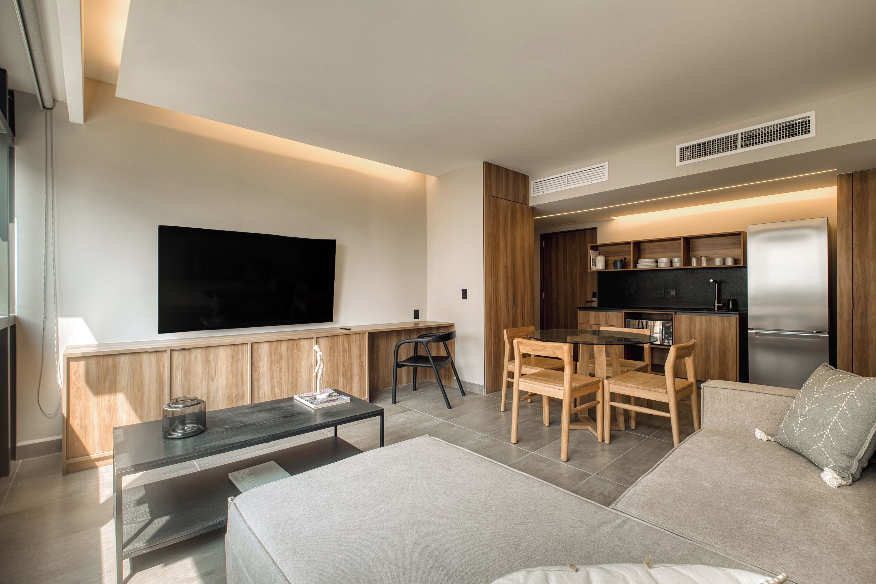 Uma sala de estar moderna com sofá, mesa de centro, televisão e cozinha, que inclui frigorífico e armários de madeira.