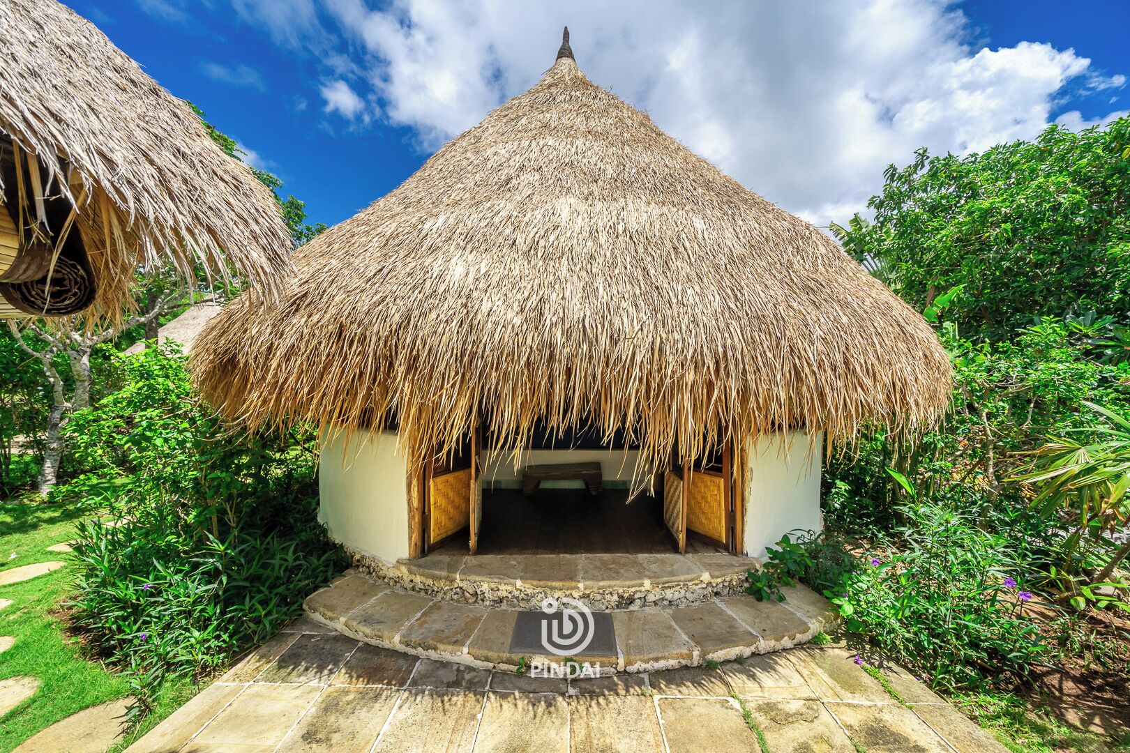 Uma cabana com telhado de palha e entrada com piso de pedra, rodeada por vegetação exuberante.