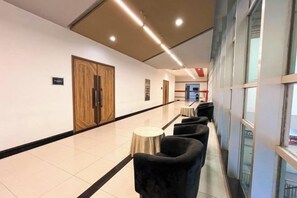 Um corredor de hotel com uma fileira de poltronas pretas, uma porta de madeira e uma pequena mesa redonda.