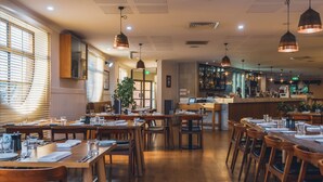 Um restaurante com mesas e cadeiras de madeira, um bar e janelas amplas com persianas.