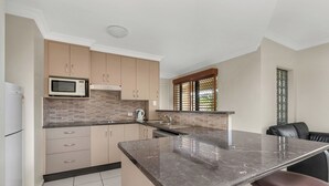 Uma cozinha com armários bege, micro-ondas, painel de azulejos, bancada de mármore, pia e janela com decoração.