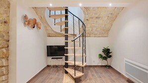 Uma escadaria em espiral com degraus de madeira e corrimãos de metal em um quarto com paredes de pedra, uma TV de tela plana e plantas em vasos.