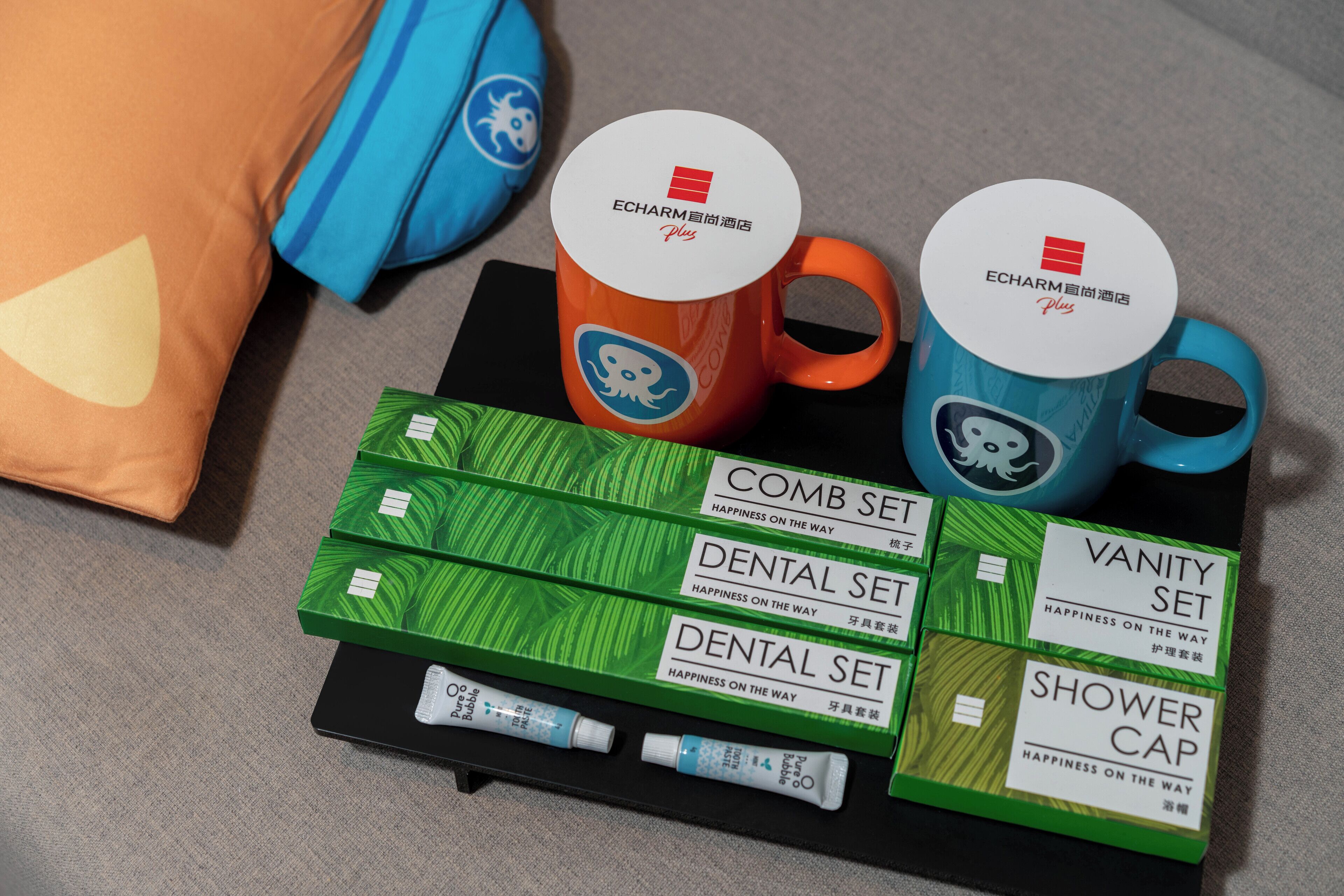 Uma bandeja com produtos de higiene bucal, incluindo pentes, kits dentários e um gorro de chuveiro, e dois copos da marca ECHARM.