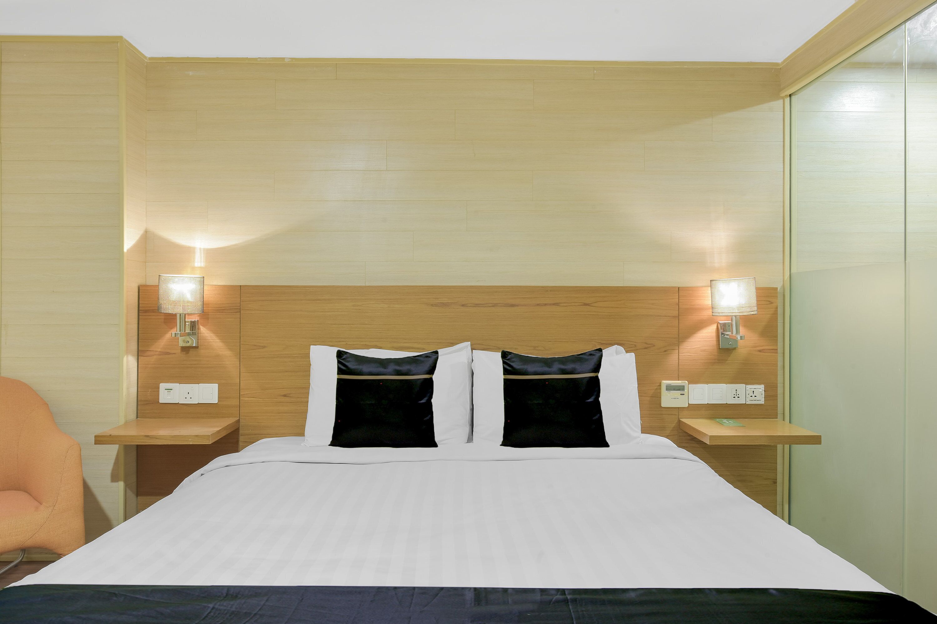 Quarto de hotel com cabeceira de madeira, duas mesas de cabeceira com luminárias, cama com roupa de cama branca e uma cadeira.