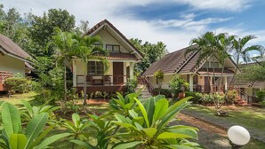 Uma casa no estilo bangalô com telhado de telhas, rodeada por vegetação exuberante e palmeiras.