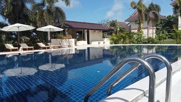 Uma piscina com água azul cristalina, rodeada por espreguiçadeiras e guarda-sóis, com palmeiras e um prédio ao fundo.