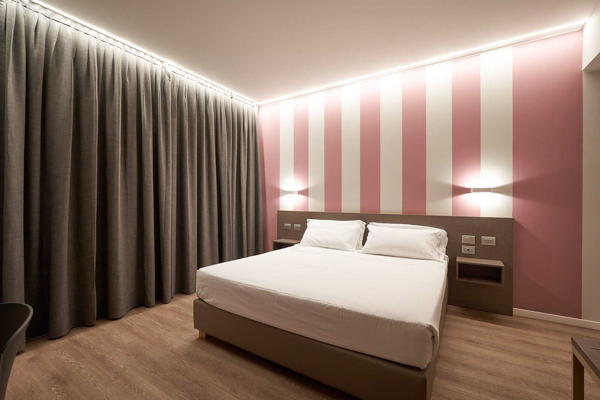 Quarto de hotel moderno com uma cama grande, uma mesinha de cabeceira e um aplique na parede.