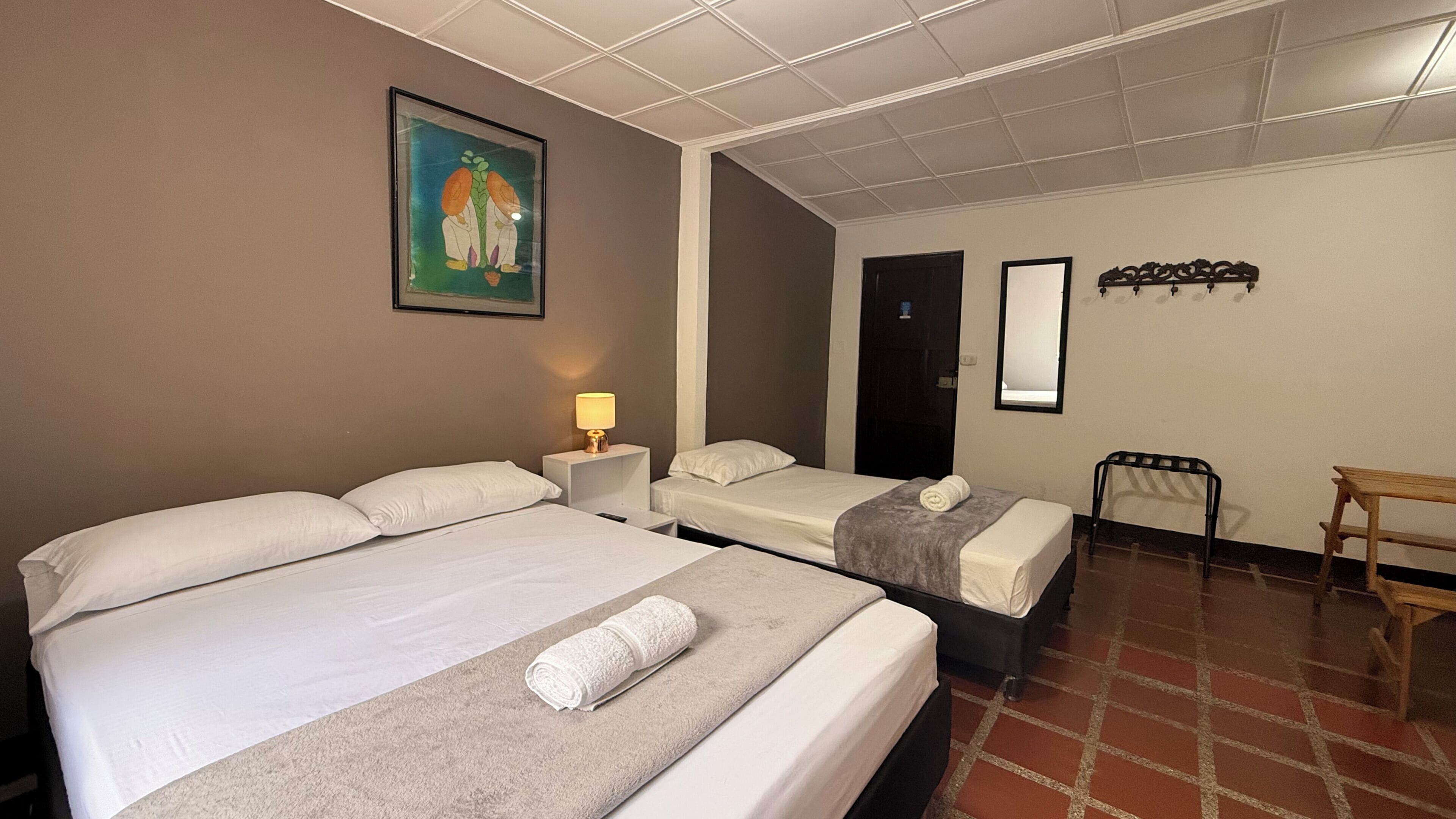 Quarto de hotel com duas camas, uma mesa de cabeceira com abajur, um espelho, um banco de madeira e um quadro na parede.