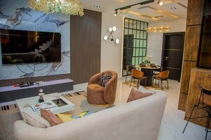 Uma sala de estar moderna com um sofá confortável, uma mesa de centro e uma TV fixada em uma parede de mármore. Há uma área de jantar com mesa e cadeiras, além de um armário de madeira.