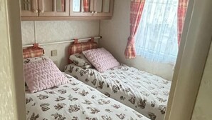 Quarto com duas camas, cada uma com roupa de cama e almofadas estampadas, um guarda-roupa de madeira e uma janela com cortina.