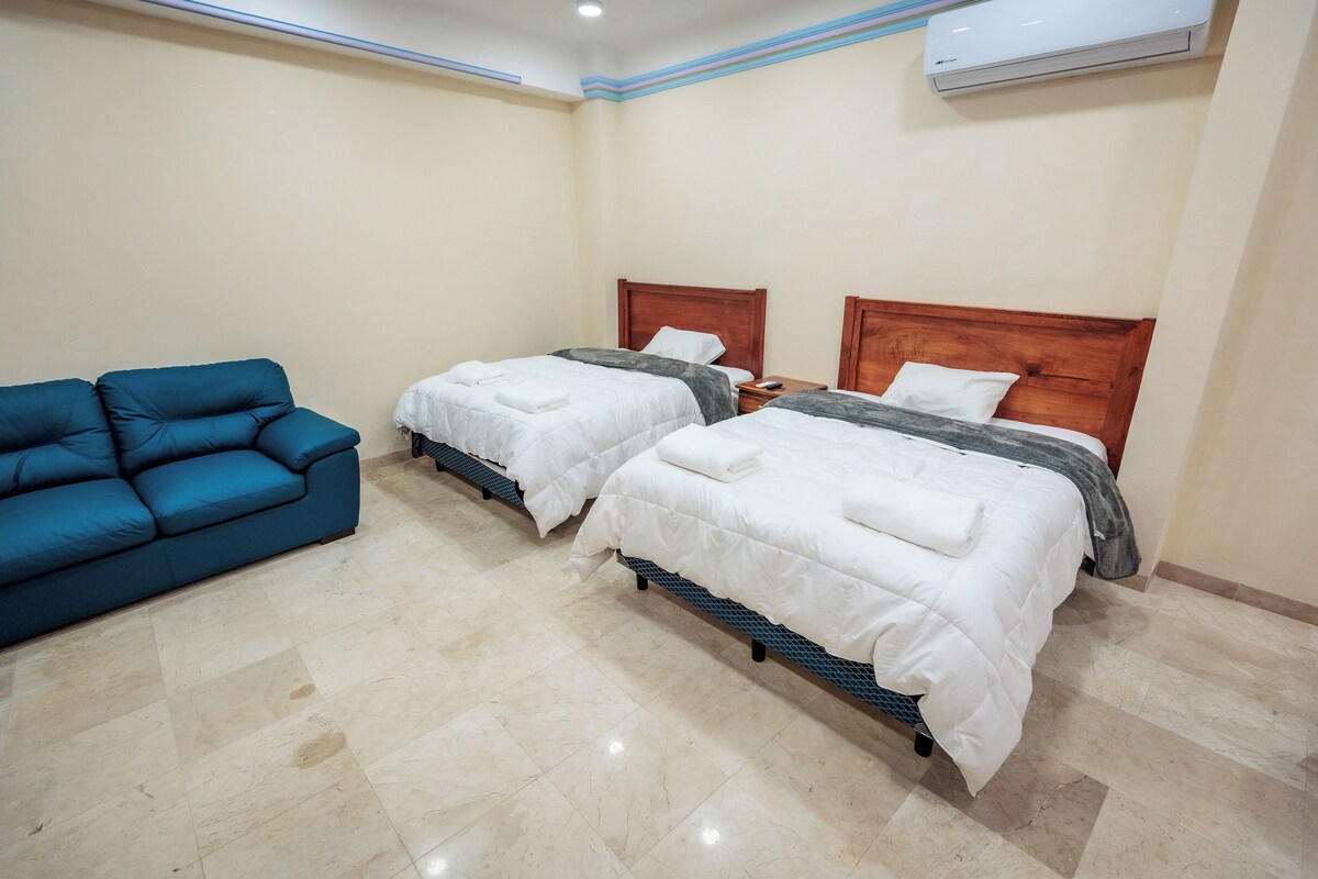Quarto de hotel com duas camas, um sofá azul e um ar condicionado na parede.