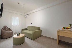 Uma sala de estar minimalista com um sofá verde, uma mesa de centro redonda, um pufe e uma fruteira sobre uma mesinha de madeira.