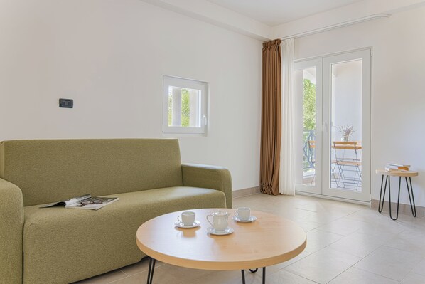 Uma sala moderna com um sofá verde, uma mesa de madeira redonda com xícaras brancas e um bule, uma janela e uma porta de vidro deslizante que dá para uma área externa.