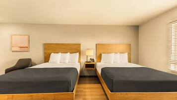 Quarto de hotel com duas camas, uma mesa de cabeceira com abajur, uma cadeira e um quadro na parede.