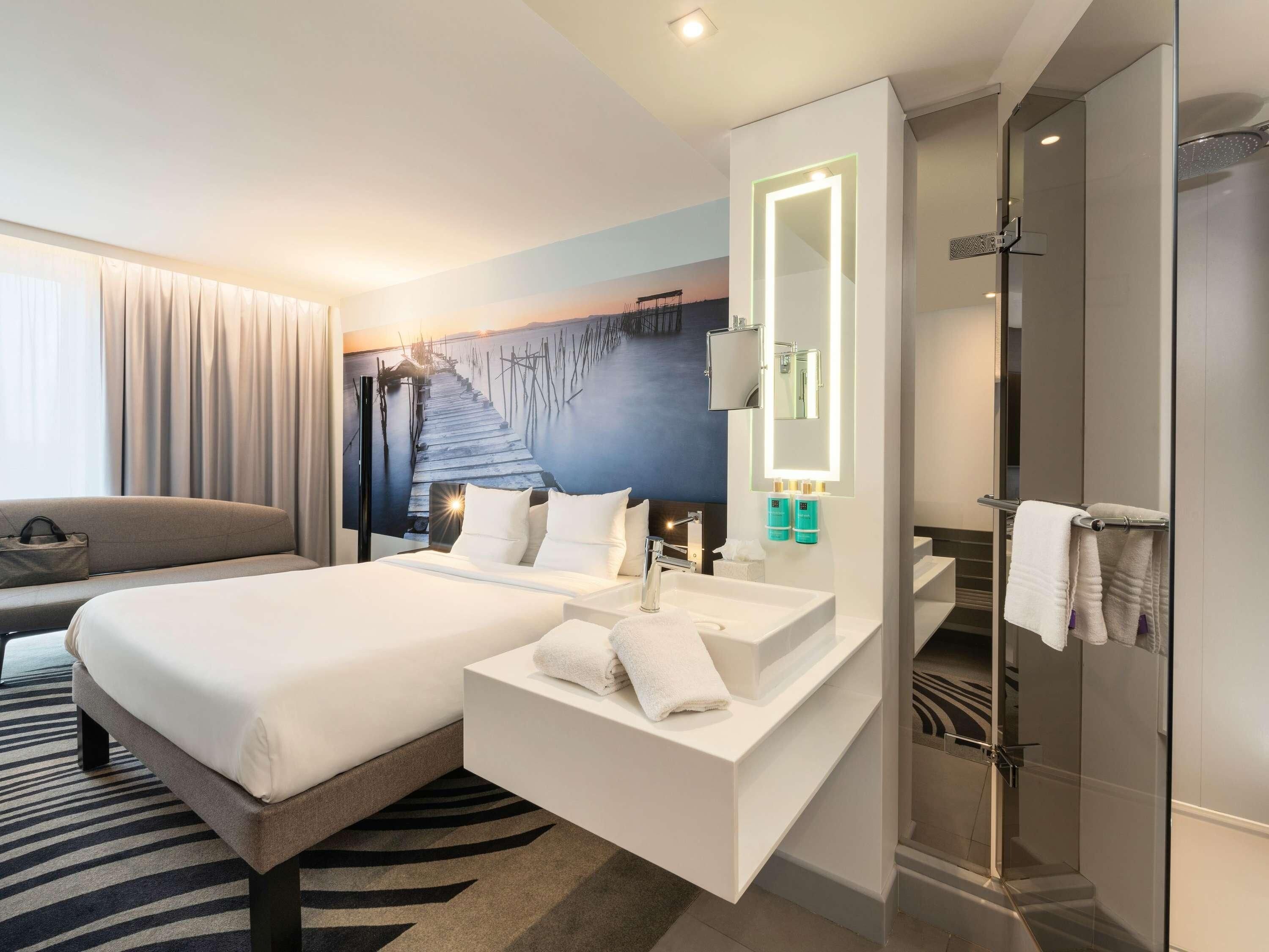 Quarto de hotel moderno com cama, banheiro com espelho e pia, e chuveiro.