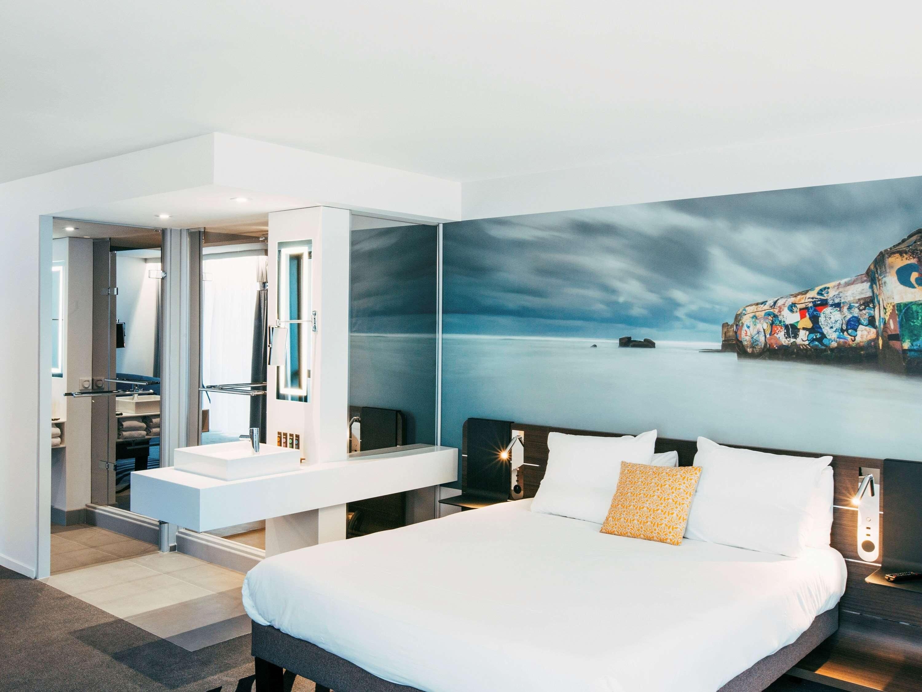 Quarto de hotel moderno com uma cama grande, banheiro com espelho e vista para o mar.