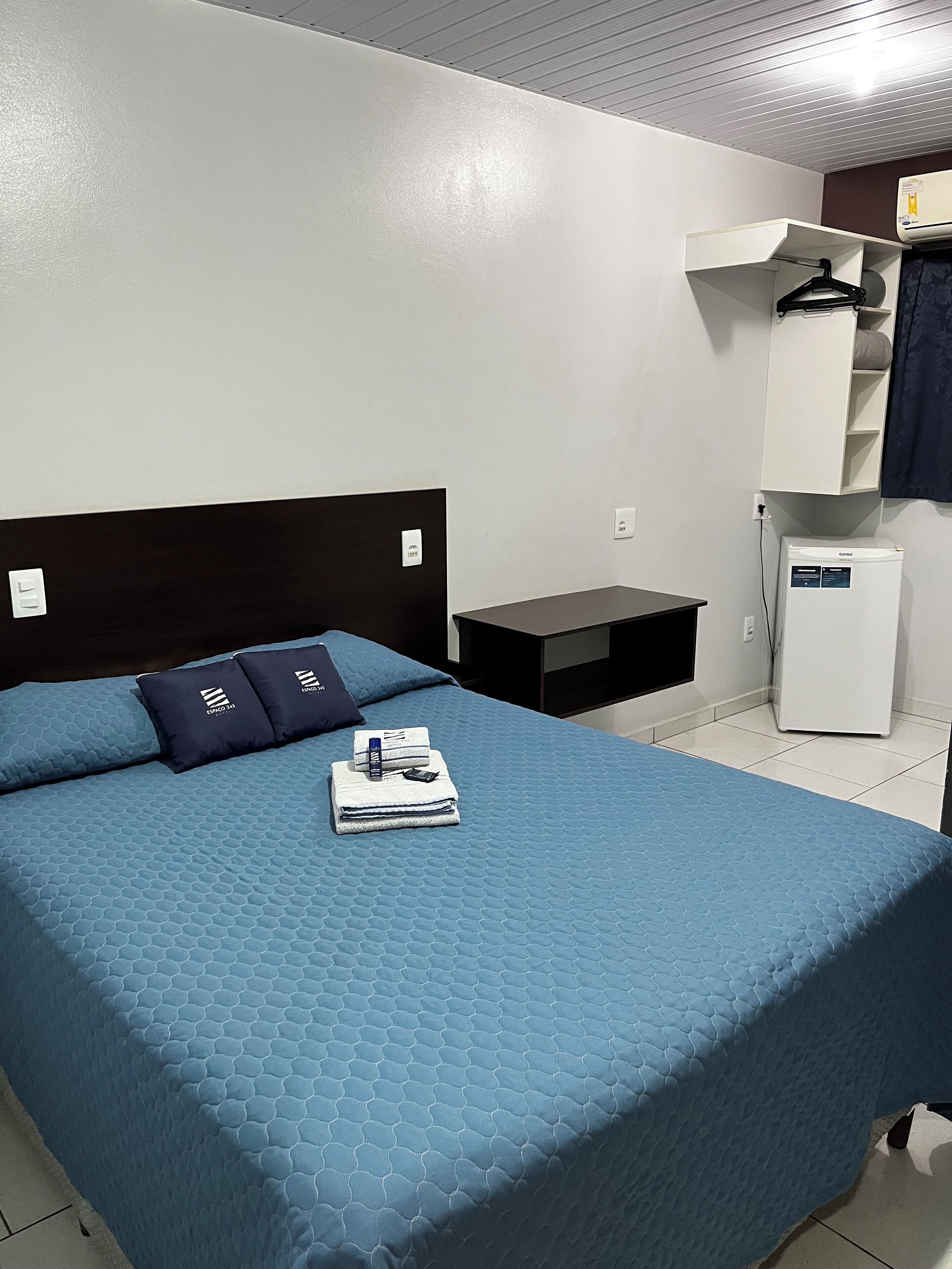 Quarto de hotel com uma cama, uma pequena escrivaninha, um guarda-roupa e um mini-frigorífico.