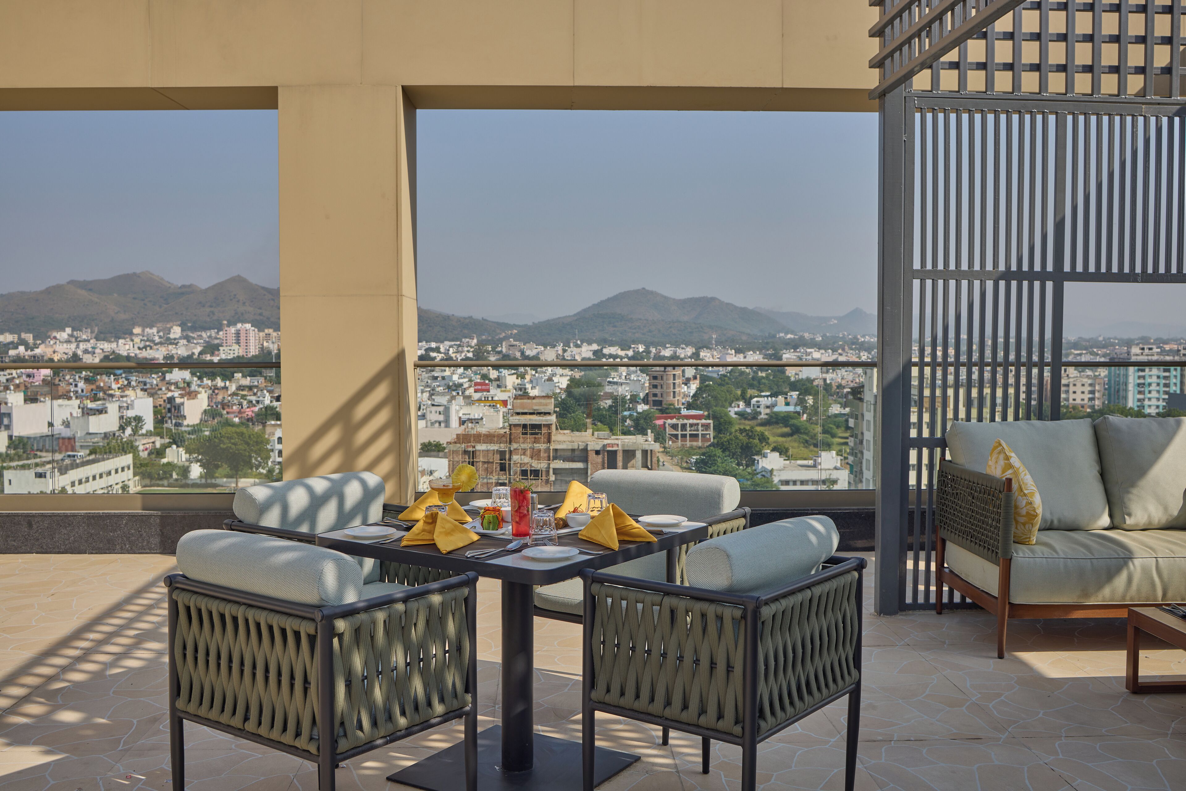 Um terraço com mobiliário de vime, uma mesa de jantar posta com guardanapos amarelos e vista para um horizonte urbano com montanhas ao fundo.
