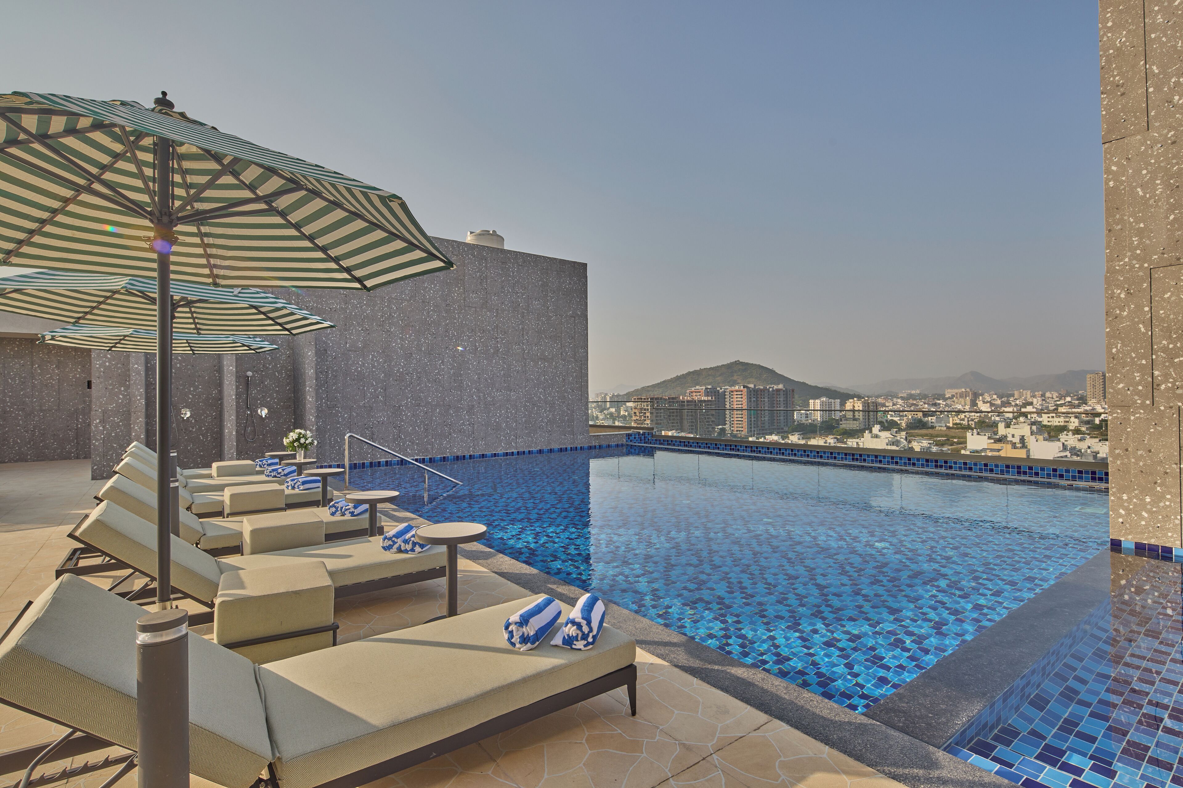 Uma área de piscina no terraço com espreguiçadeiras e guarda-sóis, piscina azulejada e vista para a cidade ao fundo.