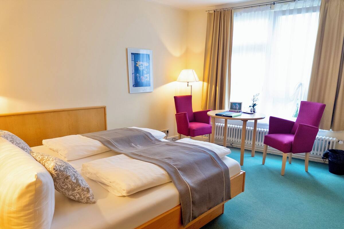 Quarto de hotel com uma cama, duas cadeiras, uma mesa, um abajur e uma janela com cortinas.