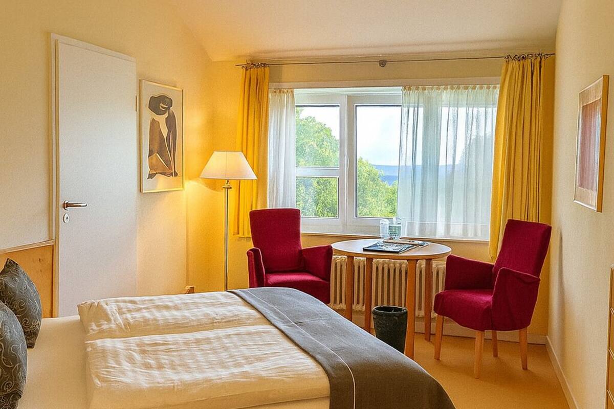Quarto de hotel com uma cama grande, duas cadeiras vermelhas, uma mesa pequena e uma janela com cortinas.