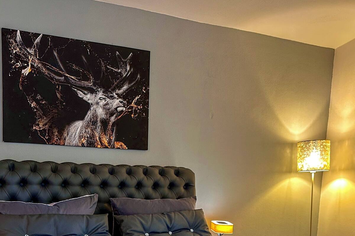 Quarto com cabeceira estofada, várias almofadas, cama com colcha cinza e azul, pintura de um veado na parede e abajur.