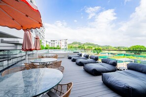 Terraço no topo do prédio com área de estar externa, incluindo mesas redondas e sofás, e guarda-corpo de vidro.