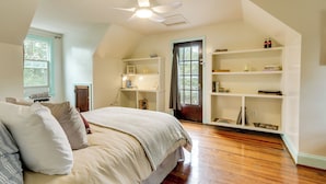 Quarto com cama grande, ventilador de teto, piso de madeira e estantes embutidas com livros e objetos decorativos.