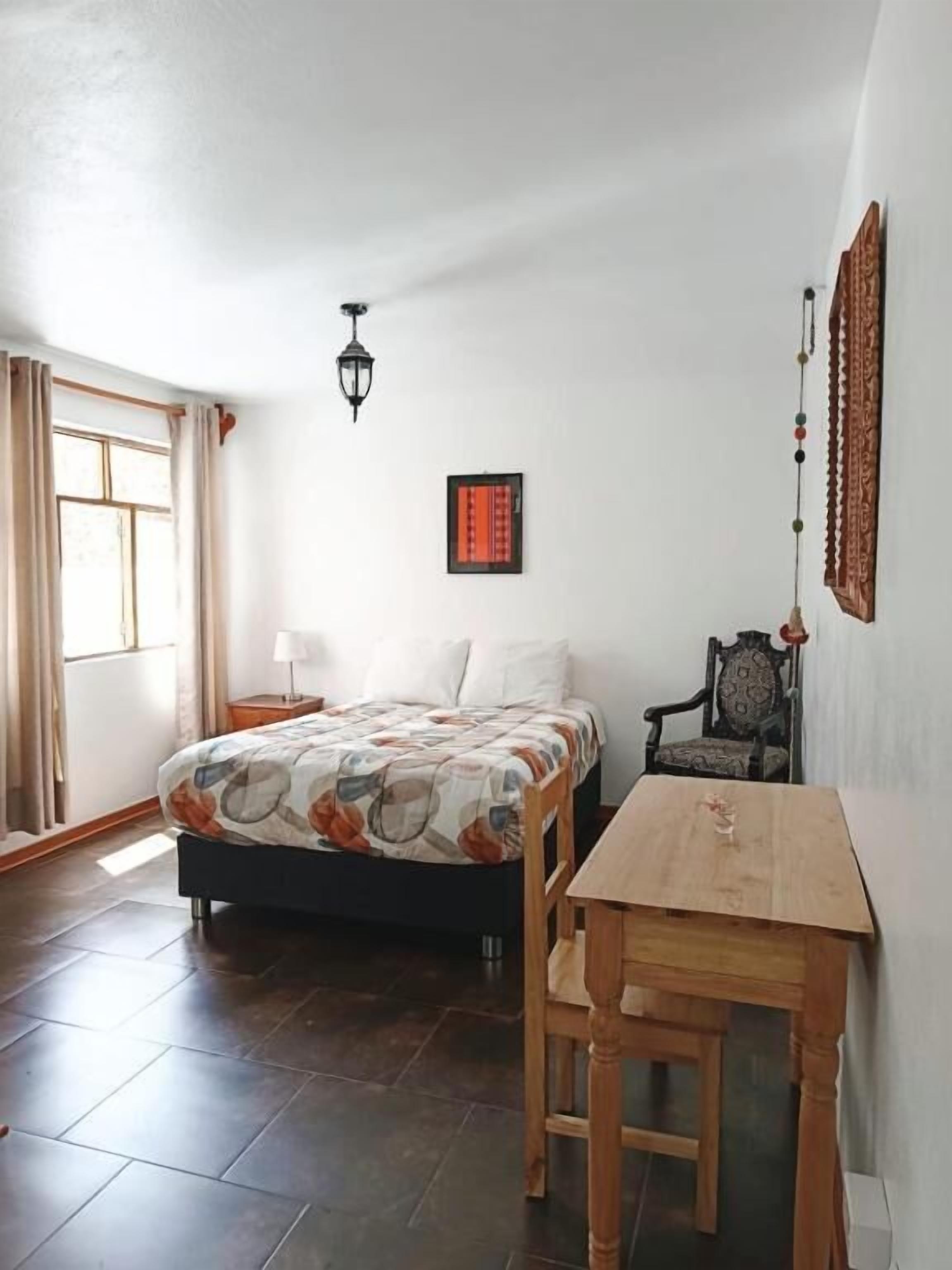 Quarto com cama, mesa de madeira, cadeira, mesa de cabeceira, abajur e decoração na parede.