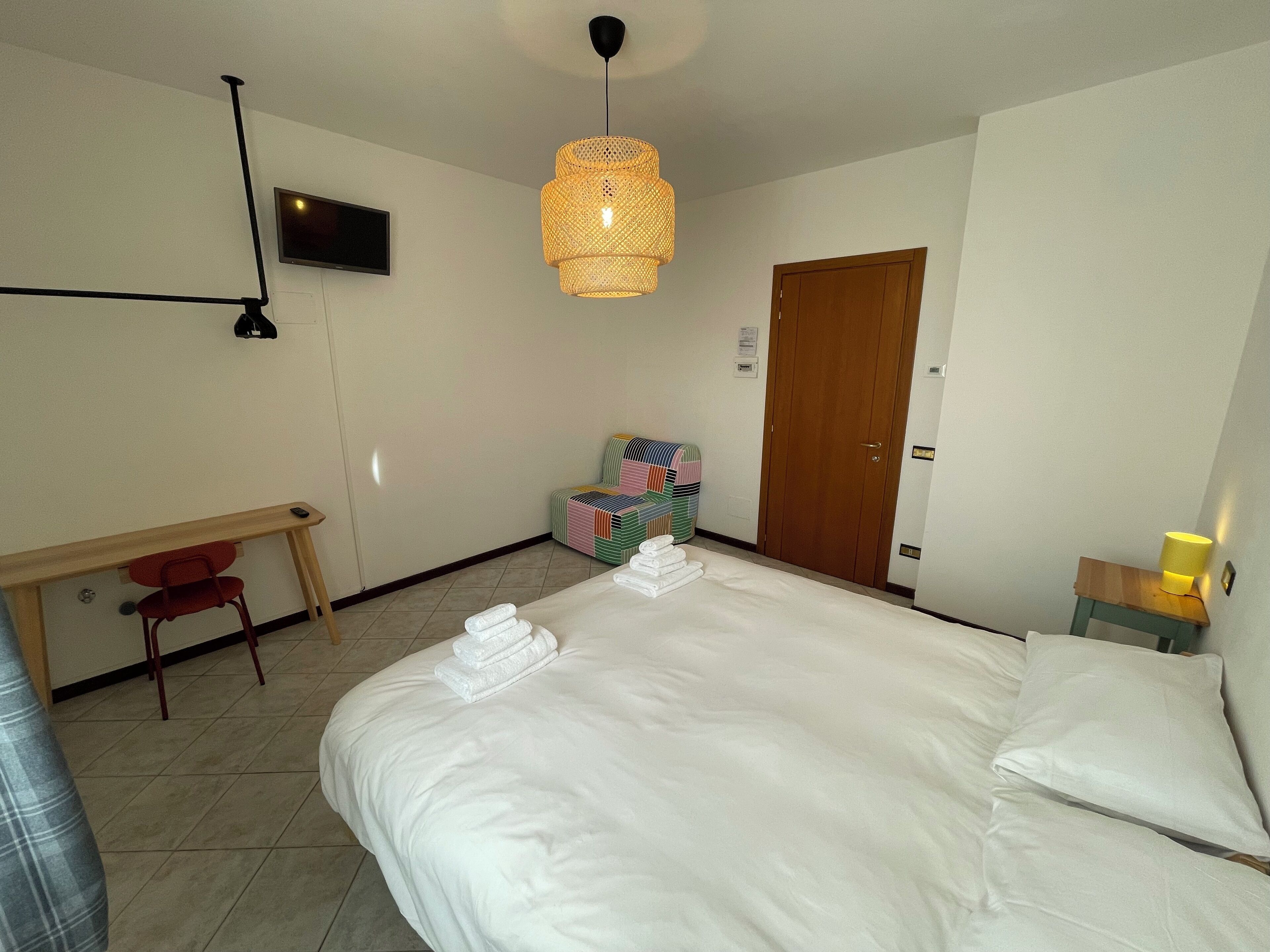 Quarto de hotel com cama, escrivaninha, cadeira, televisão, guarda-roupa e mesa de cabeceira com abajur.