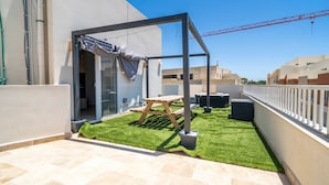 Terraço no telhado com mesa de madeira, churrasqueira e área de estar coberta.