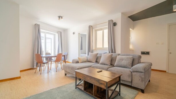 Uma sala de estar moderna com um sofá cinzento, uma mesa de centro de madeira e uma área de jantar com cadeiras.