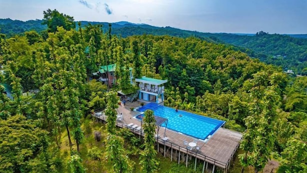 Um resort com piscina cercada por vegetação exuberante, deck de madeira e um pequeno edifício com telhado verde.