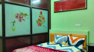 Quarto com cama, almofadas coloridas, cabeceira de madeira, parede verde e um objeto decorativo vermelho sobre a cama.