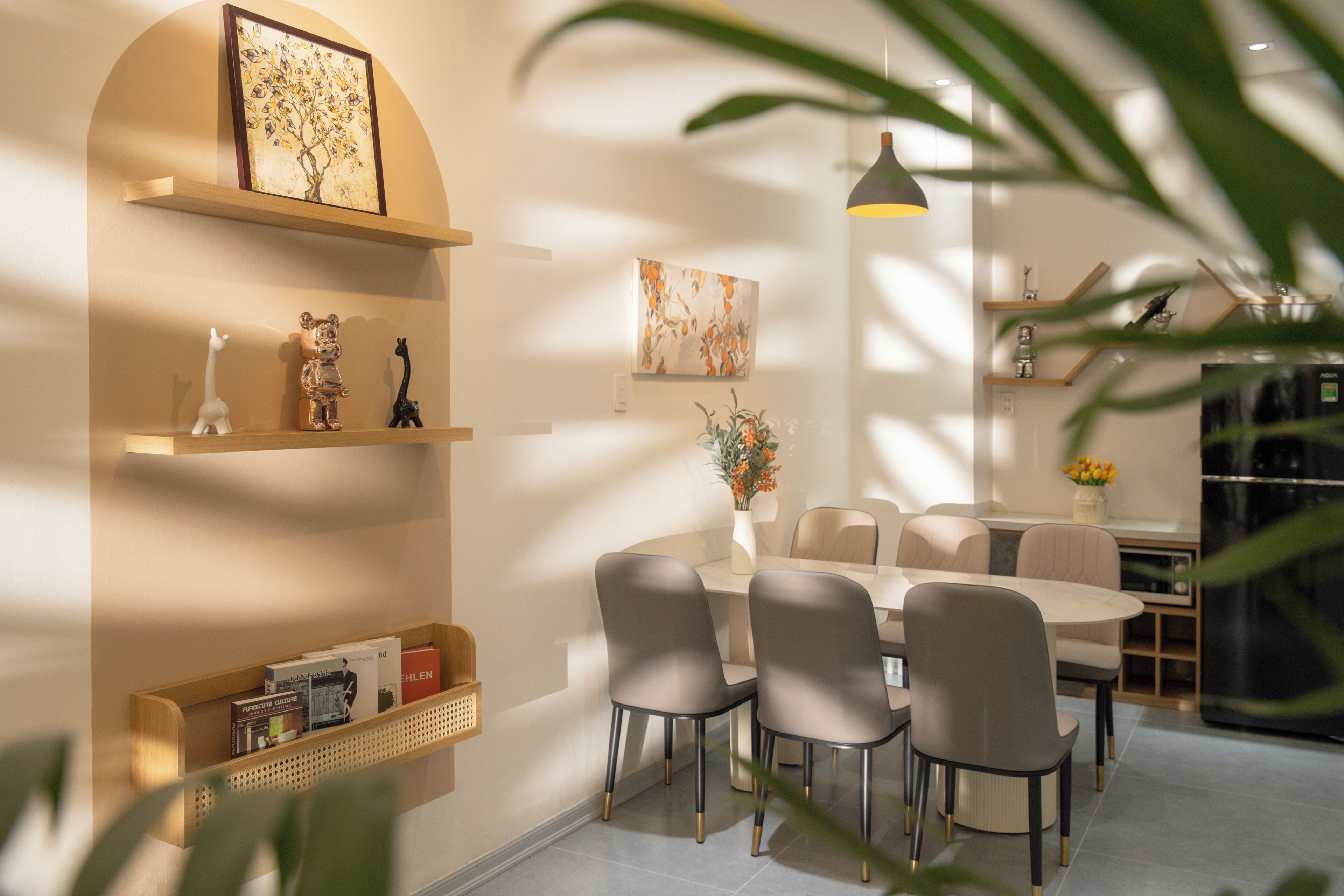 Uma sala de jantar moderna com uma mesa redonda, seis cadeiras e uma luminária pendente. Há prateleiras com objetos decorativos e um vaso com flores na parede.