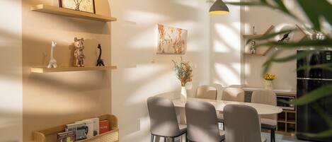 Uma sala de jantar moderna com uma mesa redonda, seis cadeiras e uma luminária pendente. Há prateleiras com objetos decorativos e um vaso com flores na parede.