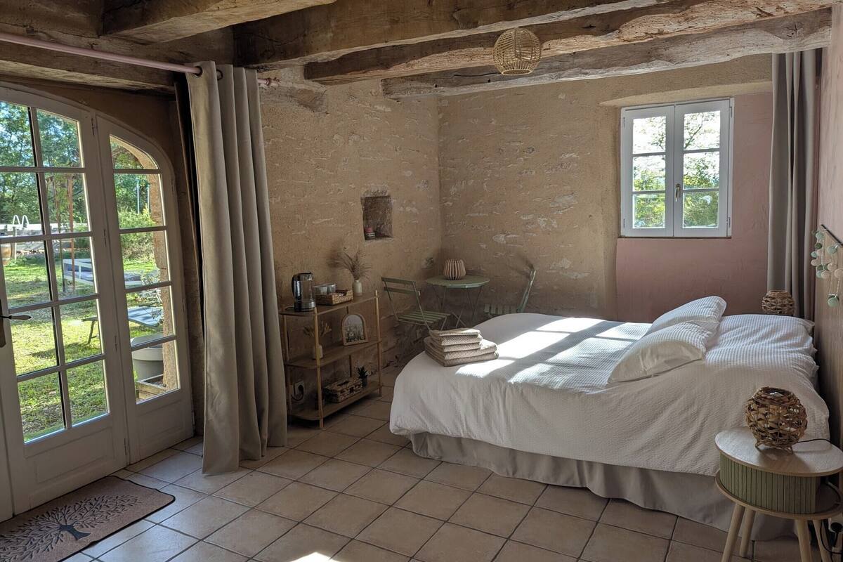 Quarto rústico com cama, uma mesinha, cadeira, janela e porta que dá para uma área externa.