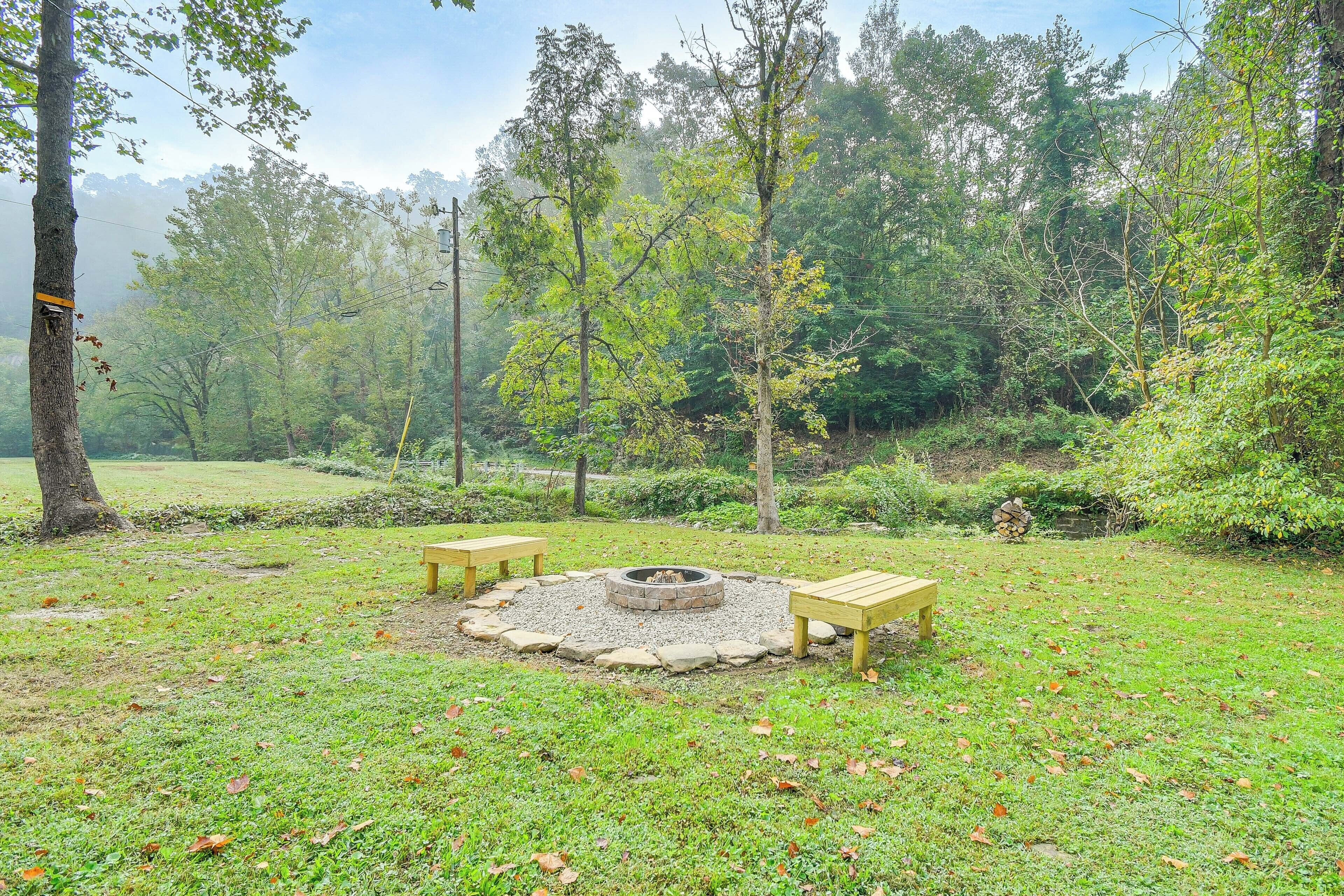 Uma fogueira circular com bancos, cercada por uma borda de pedra, situada em uma área gramada com árvores ao fundo.